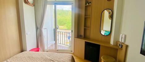 Quarto com cama, um armário de madeira, espelho, cadeira vermelha e varanda com vista para o verde.