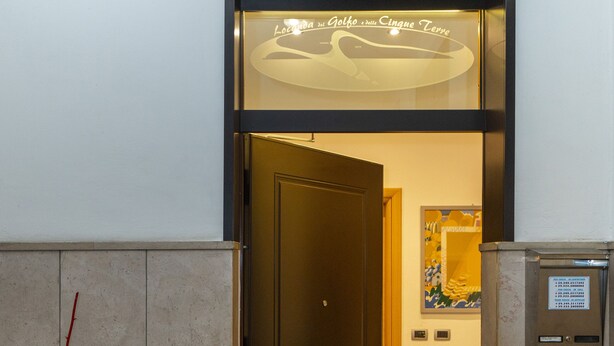 Entrada de hotel com porta de madeira escura, piso revestido e tapete de boas-vindas, além de placa na parede.