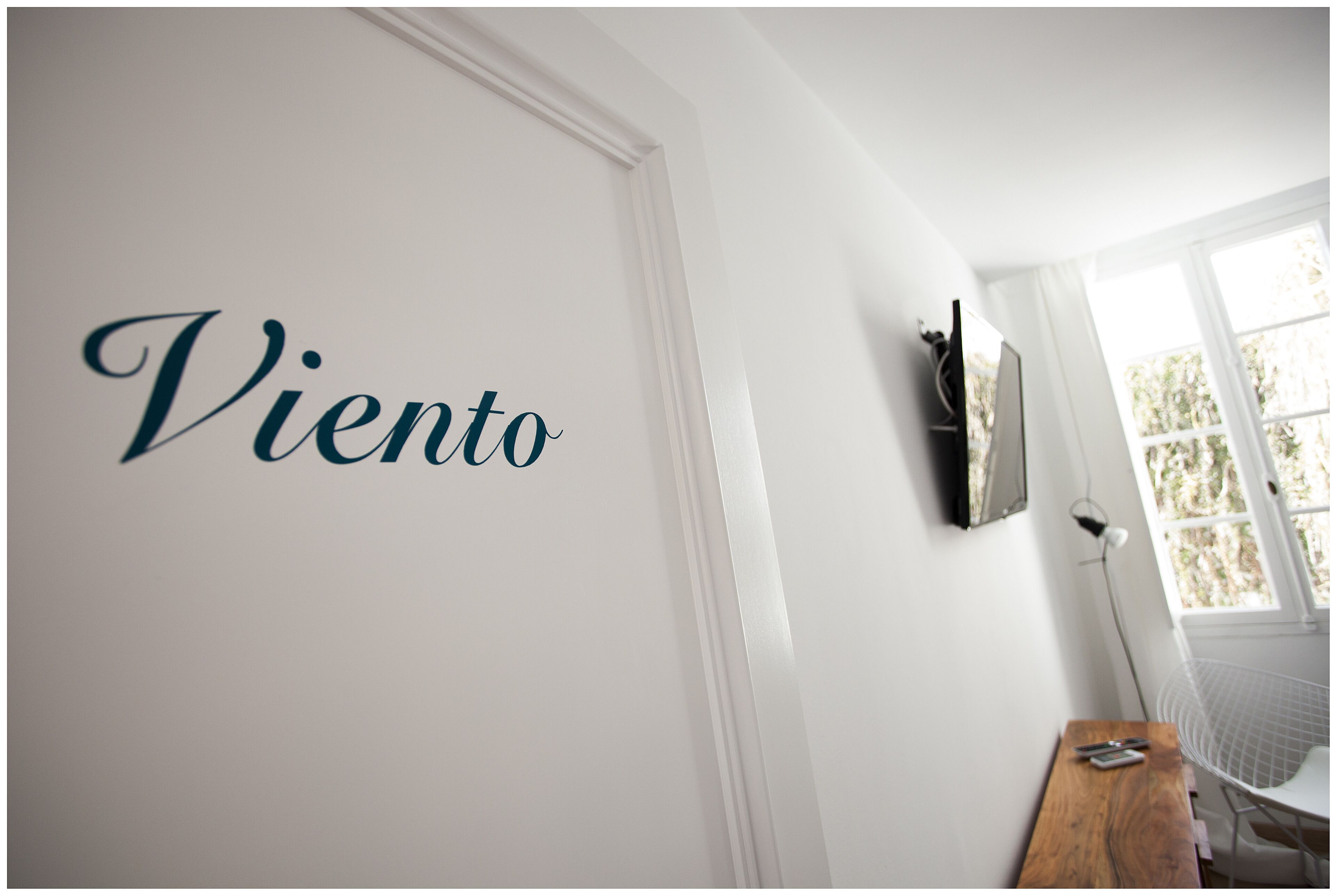 Quarto com parede branca exibindo a palavra "Vienno" em caligrafia. Há uma mesa de madeira com uma cadeira, um abajur e uma janela com vista para o verde.