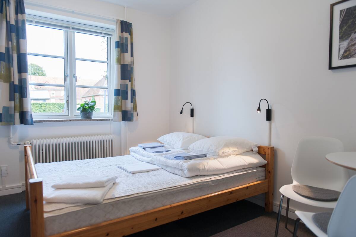 Cama bem arrumada com lençóis brancos, mesa de cabeceira, cadeira, radiador e janela com cortinas.