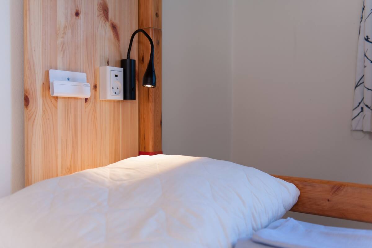Uma cama bem arrumada com roupa de cama branca, cabeceira de madeira, uma tomada na parede e uma luminária preta.