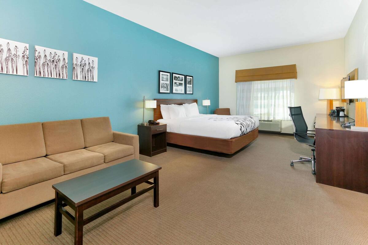 Quarto de hotel com cama, sofá, escrivaninha e mesa de centro. Há quadros nas paredes e uma janela com cortinas.