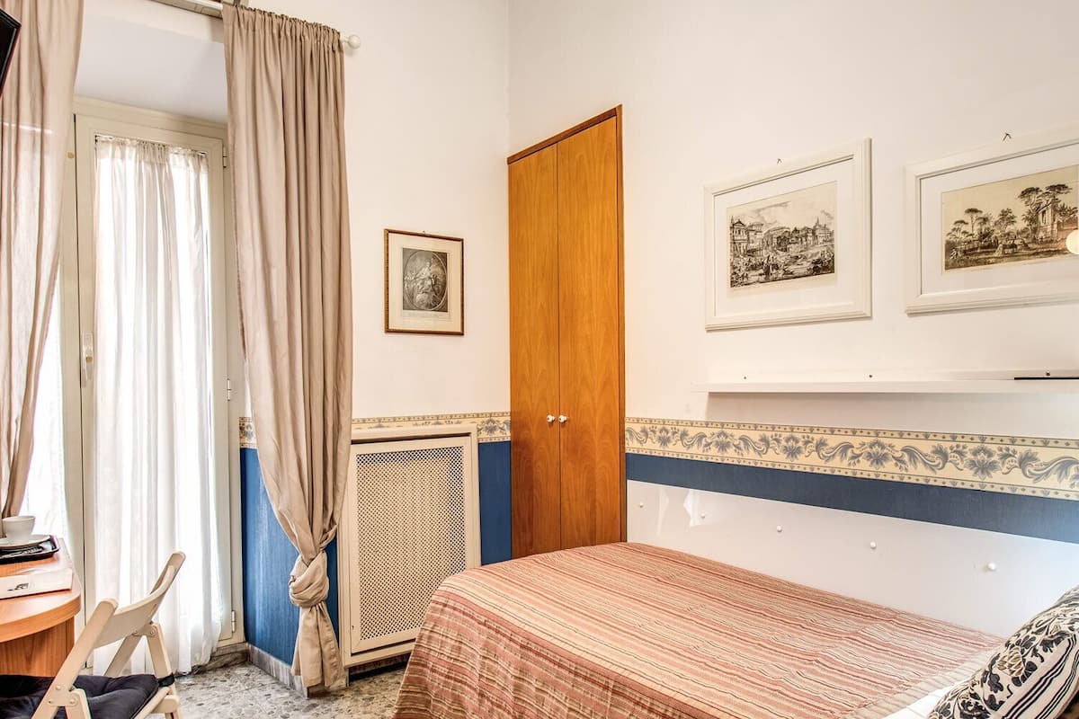 Quarto de hotel com uma cama, uma escrivaninha com computador, uma cadeira, um radiador e duas imagens emolduradas na parede.