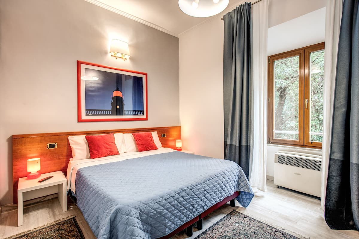 Quarto de hotel com uma cama grande, mesa de cabeceira com luminária, quadro na parede, janela com cortinas e radiador.