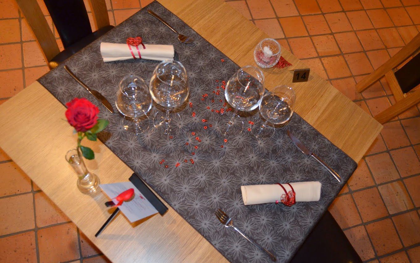 Uma mesa de jantar com toalha de mesa cinza estampada, uma rosa vermelha e vários copos de vinho e guardanapos.
