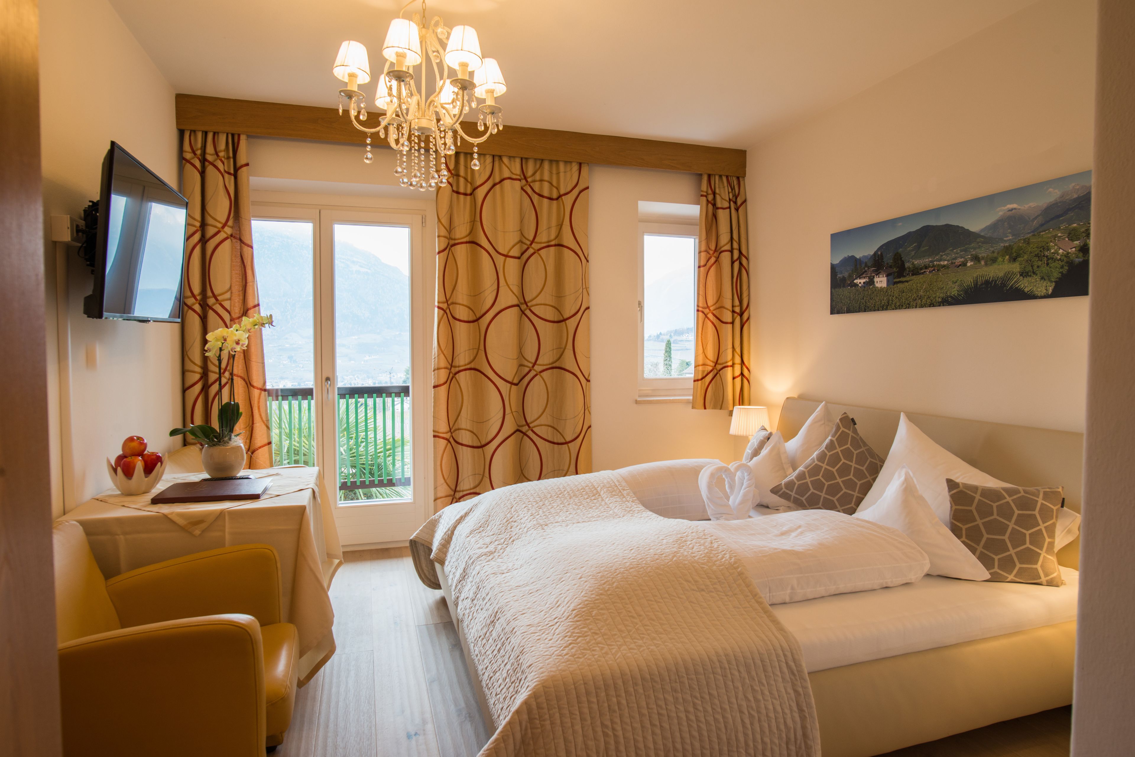 Quarto de hotel com cama, televisão, mesa de jantar com cadeiras, janela com cortinas e um quadro na parede.