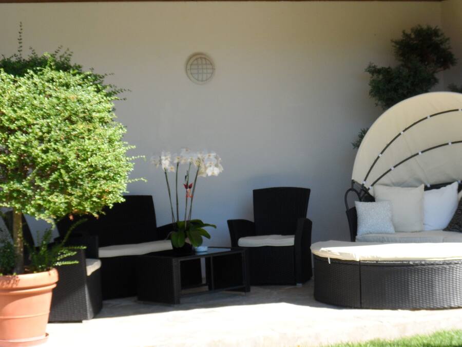 Área de pátio com móveis de exterior, incluindo um sofá redondo de vime com almofadas, duas cadeiras pretas e uma mesa pequena. Há uma planta em vaso e um conjunto de flores brancas sobre um suporte.