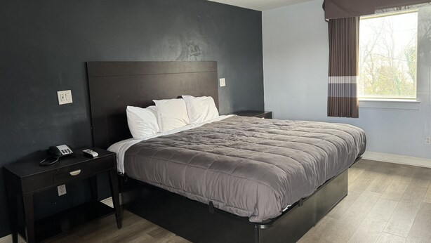 Um quarto moderno com cabeceira escura, uma cama com colcha cinza, uma mesa de cabeceira com um telefone e uma janela com cortinas.