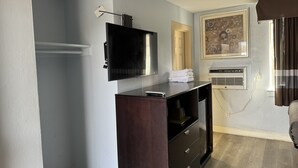 Quarto de hotel com televisão de tela plana na parede, cômoda de madeira escura com gavetas, uma obra de arte emoldurada sobre a cômoda e uma janela com cortinas.