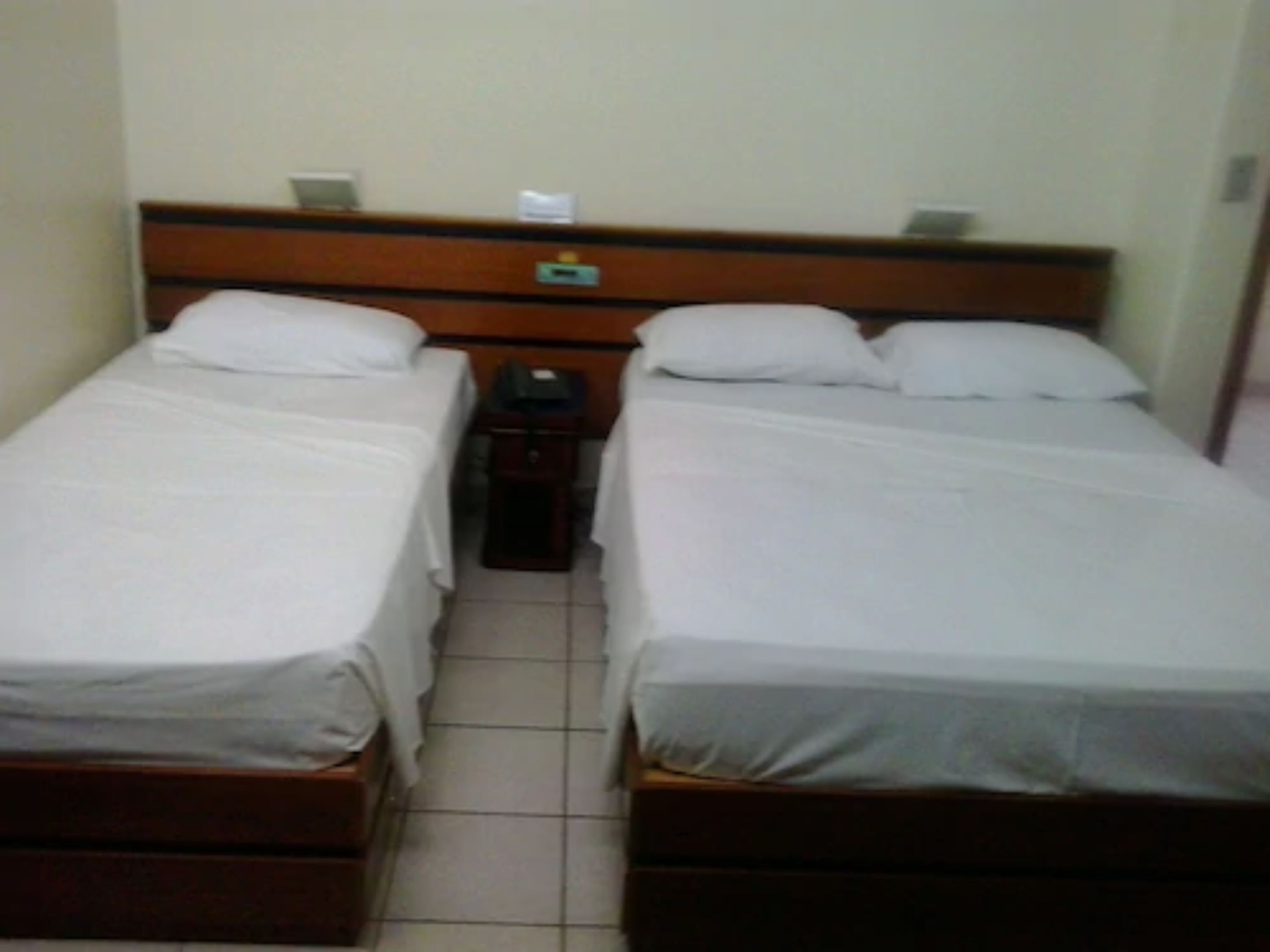 Quarto de hotel com duas camas de solteiro, cabeceira de madeira, mesa de cabeceira com abajur e uma pequena mesa.