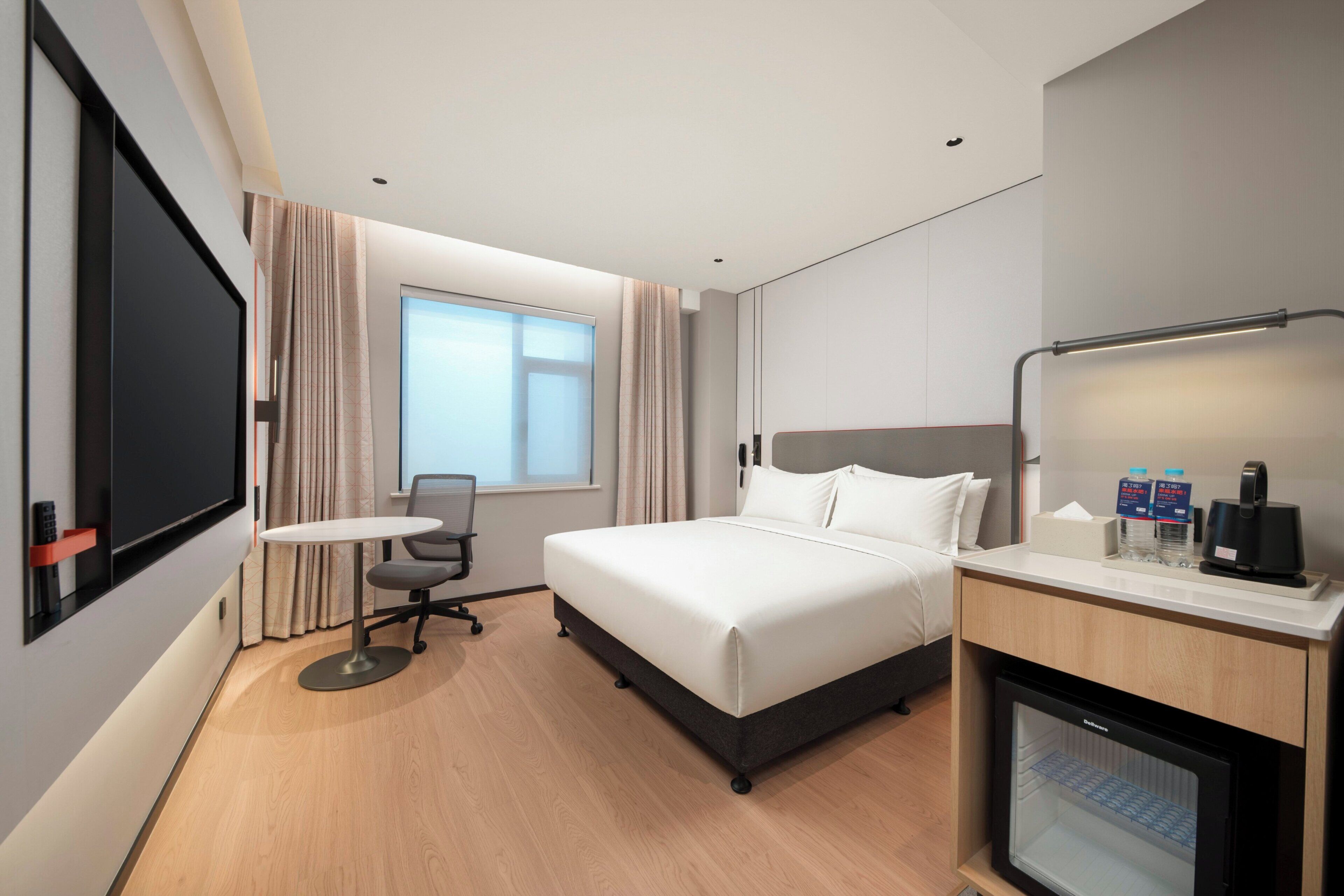 Quarto de hotel com cama, escrivaninha, cadeira, televisão, frigobar e janela com cortinas.