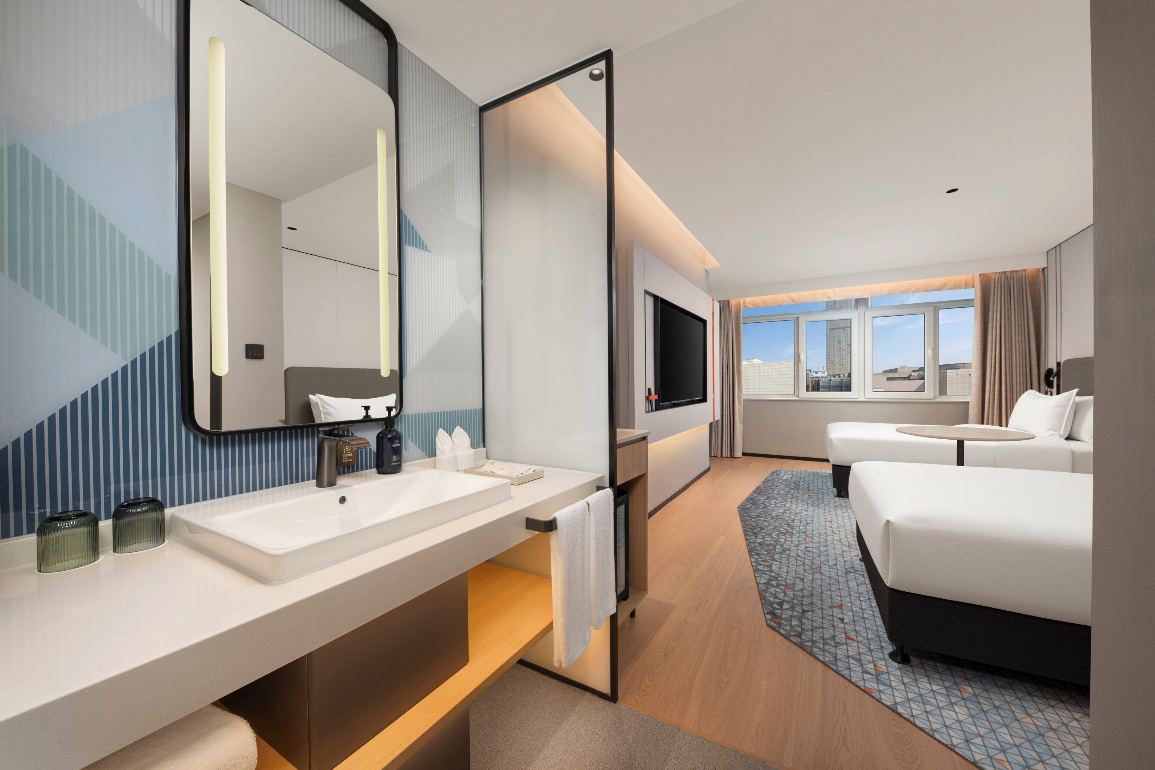 Quarto de hotel moderno com um espelho grande, pia branca, televisão de tela plana, cama e janela com vista para a cidade.