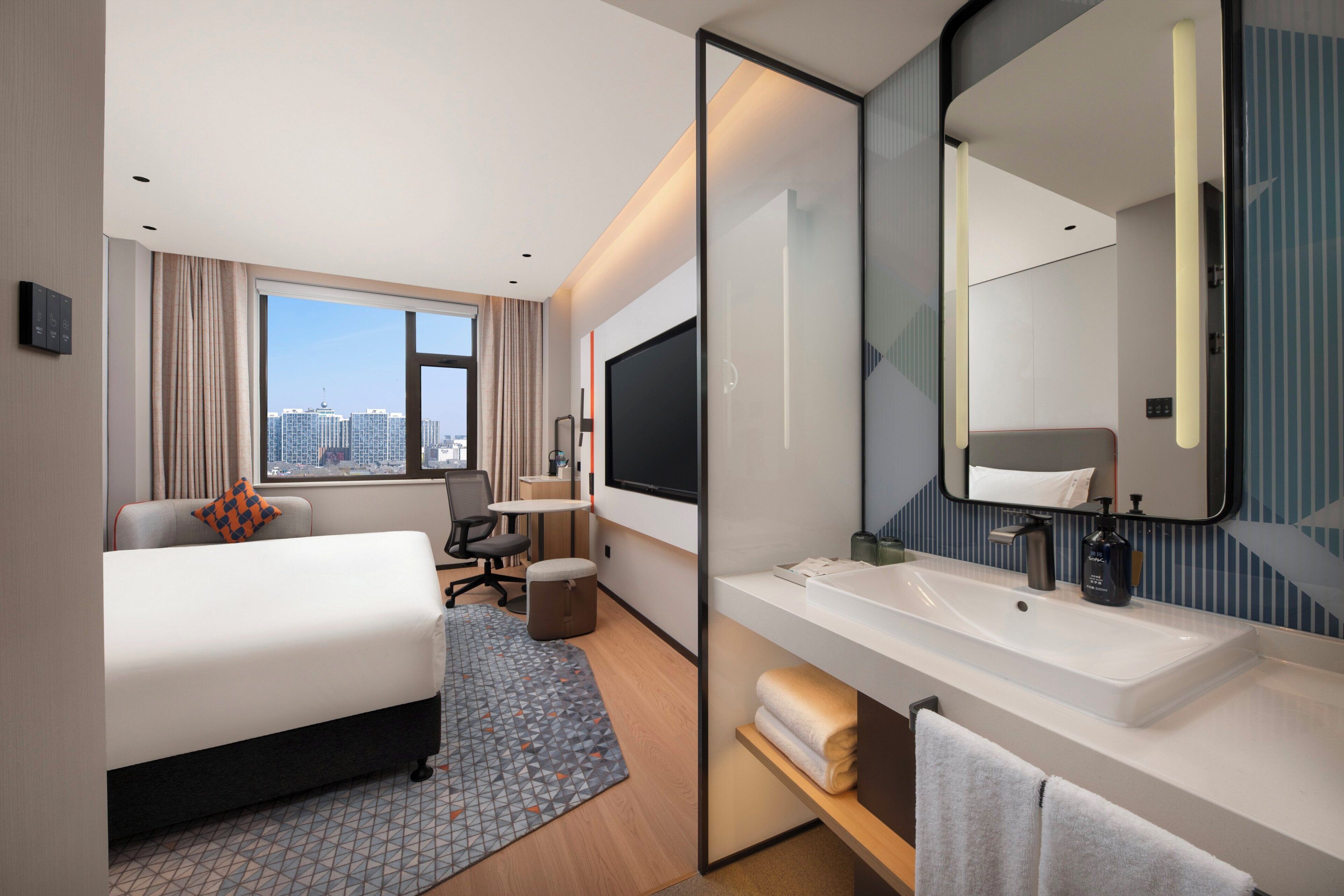 Quarto de hotel com cama, escrivaninha, cadeira e uma janela grande com vista para a cidade.