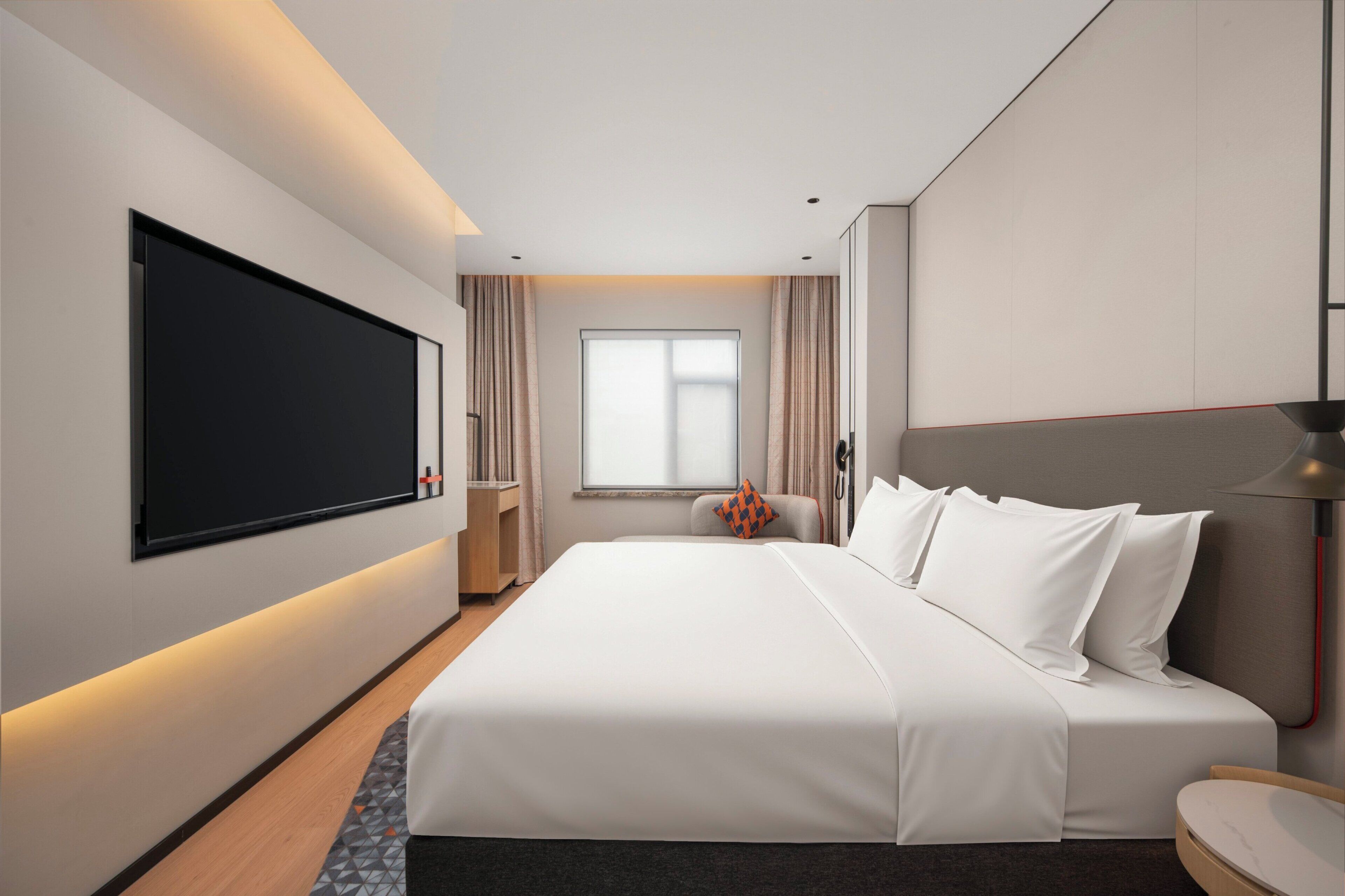Quarto de hotel moderno com uma cama grande, televisão de tela plana, mesa de cabeceira e janela com cortinas.