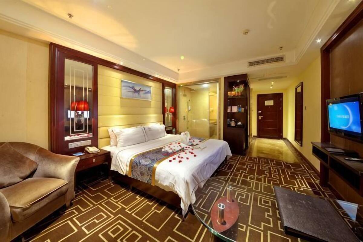 Quarto de hotel com cama, uma cadeira, televisão e escrivaninha com abajur.
