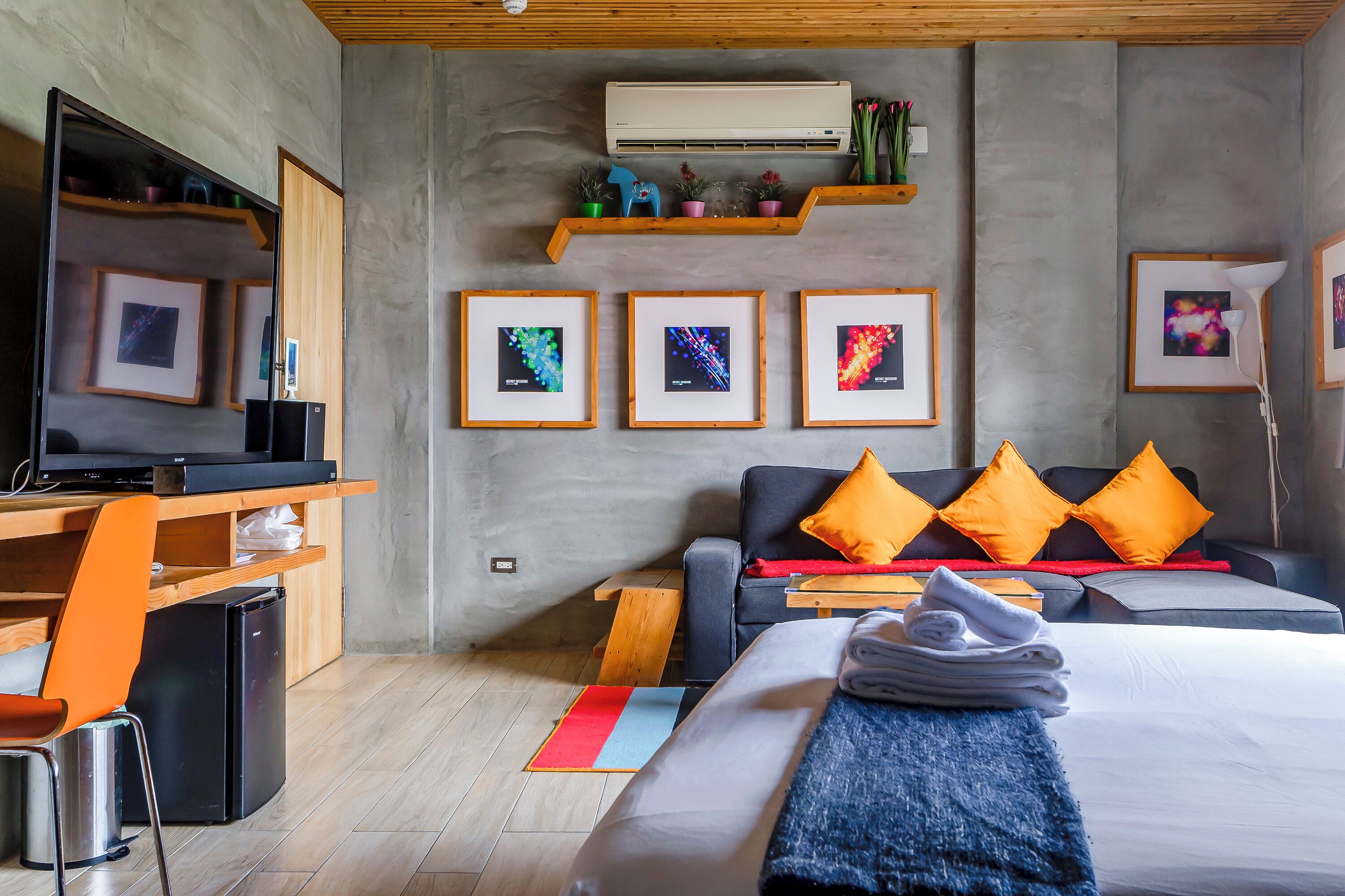 Quarto de hotel moderno com uma cama, uma escrivaninha com computador, um sofá com almofadas laranja e obras de arte emolduradas na parede.