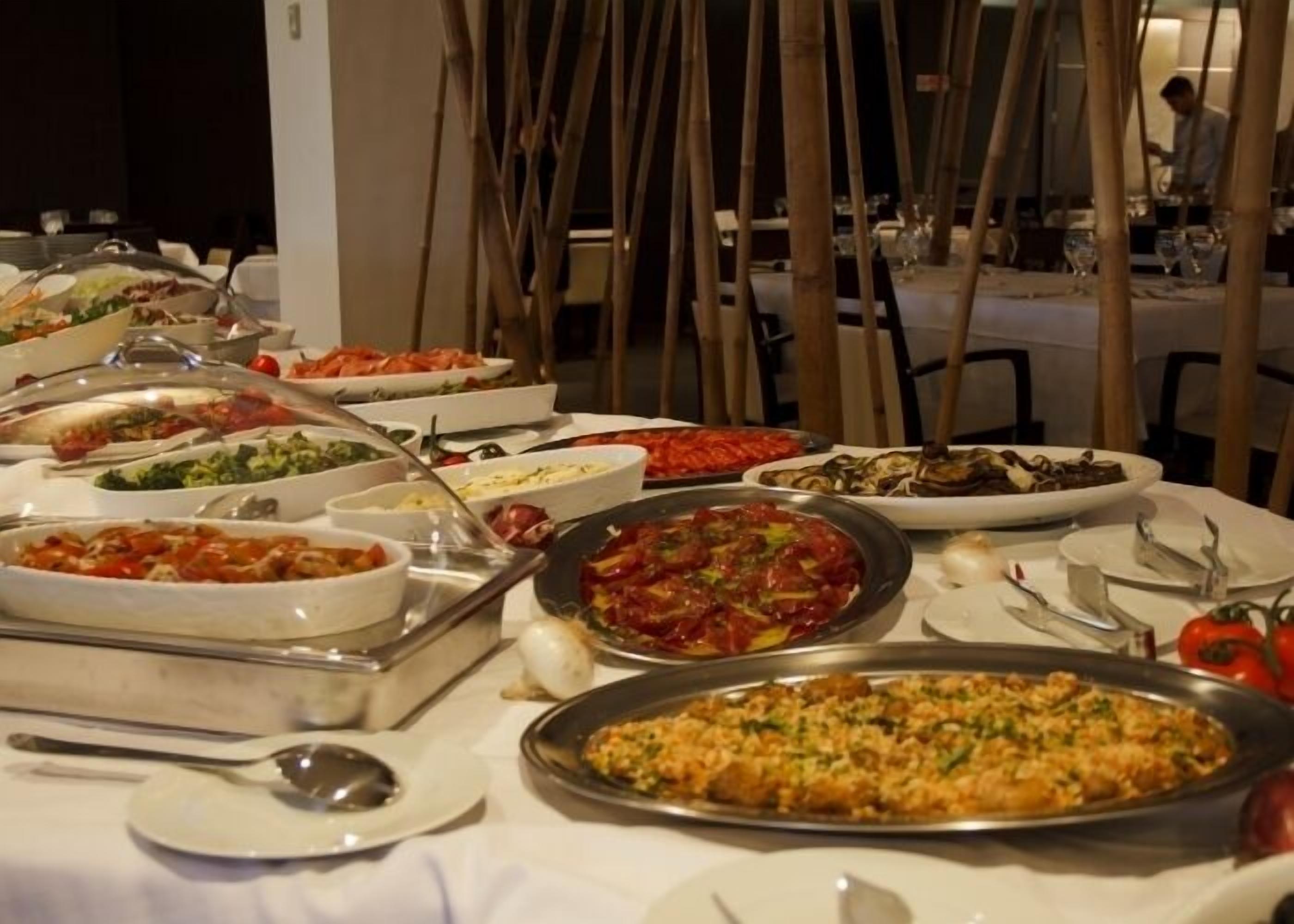 Uma mesa de buffet com diversos pratos, incluindo massas, saladas e outros alimentos prontos, montada em uma área de refeições com cadeiras e mesas.