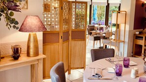 Uma área de jantar com uma mesa posta para dois, um abajur decorativo e vista para a área externa através de um painel de madeira.