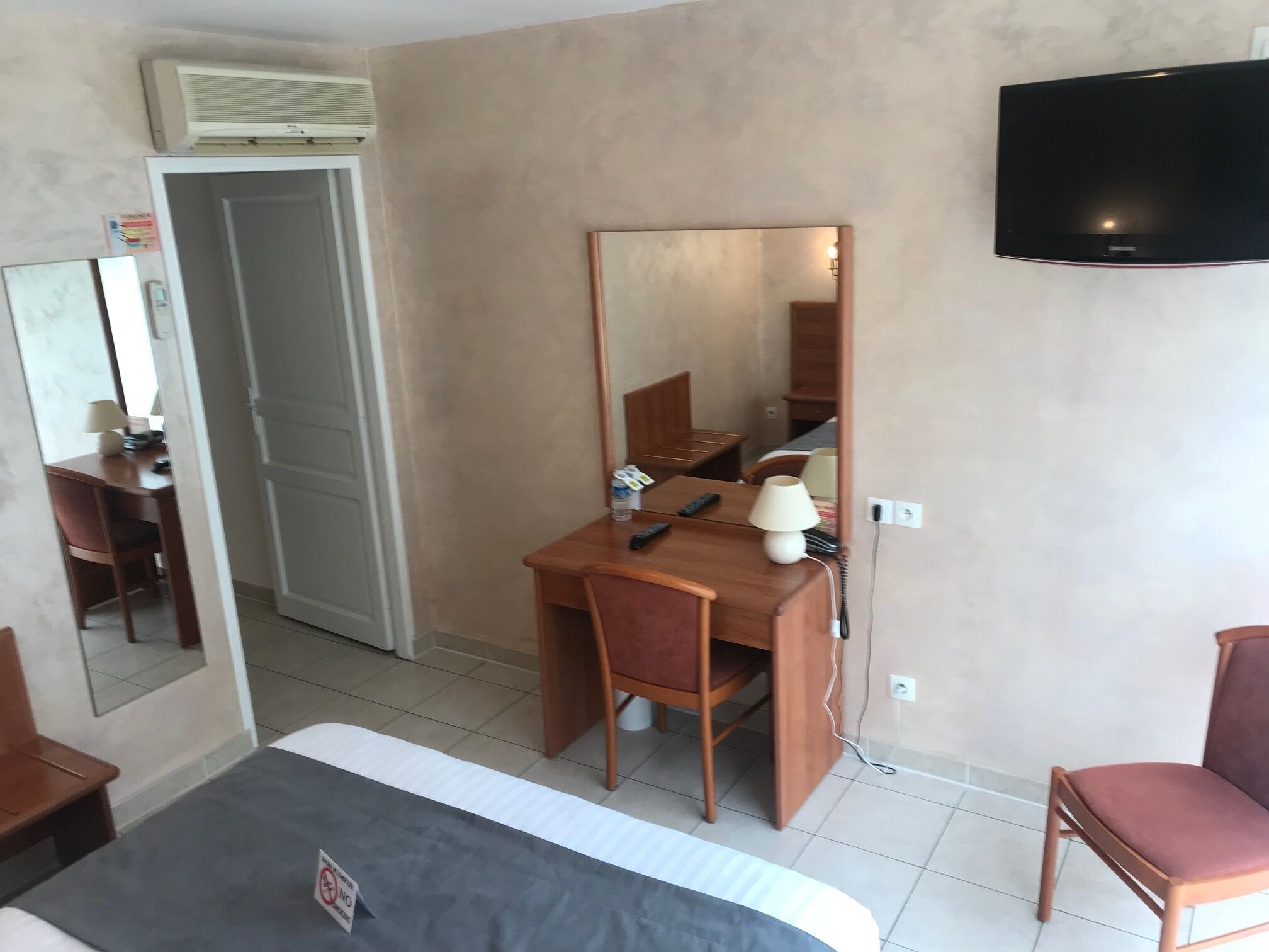 Quarto de hotel com cama, escrivaninha, cadeira, espelho, televisão e ar condicionado.