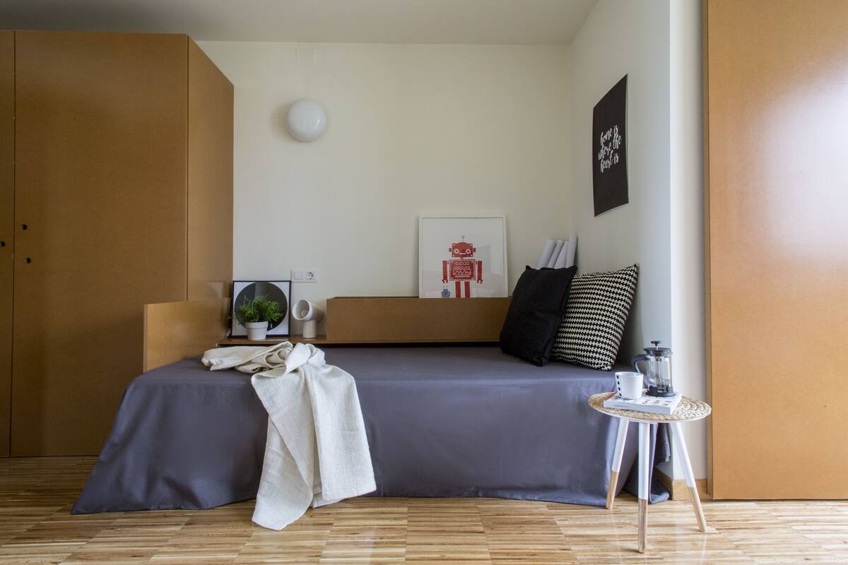 Quarto moderno com cama, mesa de cabeceira e uma pequena mesa redonda com uma xícara e um livro.