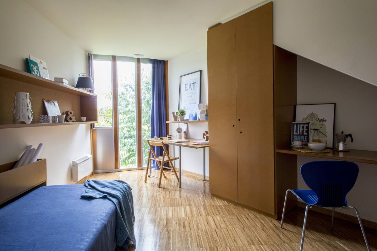 Quarto moderno com cama, escrivaninha e cadeira. Há uma janela com vista para o verde lá fora.