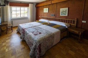 Uma cama de casal com cabeceira de madeira, duas mesas de cabeceira, uma escrivaninha com cadeira, um radiador e uma janela com cortinas.