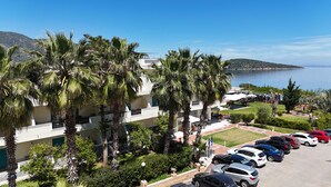 Um hotel com vários andares, um estacionamento com diversos carros e vista para o mar e colinas ao fundo.