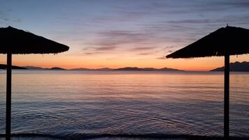 Cena de praia com dois guarda-sóis de palha e água calma refletindo o pôr do sol.