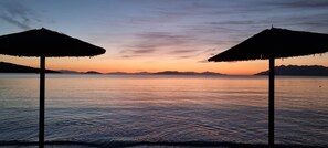 Cena de praia com dois guarda-sóis de palha e água calma refletindo o pôr do sol.