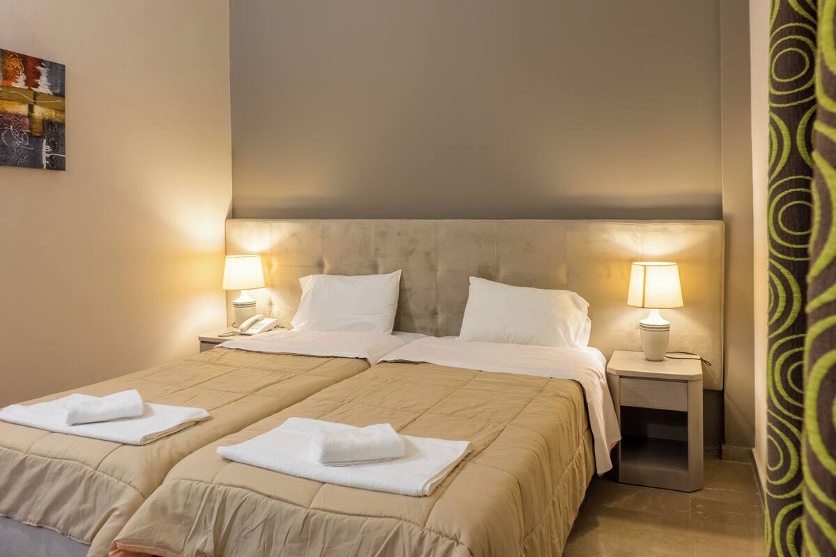 Quarto de hotel com duas camas, cada uma com colcha bege e almofadas brancas. Há luminárias de cabeceira e um telefone nas mesas de apoio.