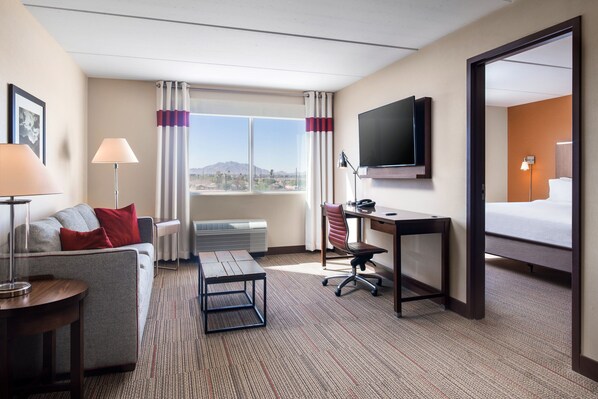 Quarto de hotel com cama, escrivaninha, cadeira, televisão, abajur, sofá e janela com cortinas.