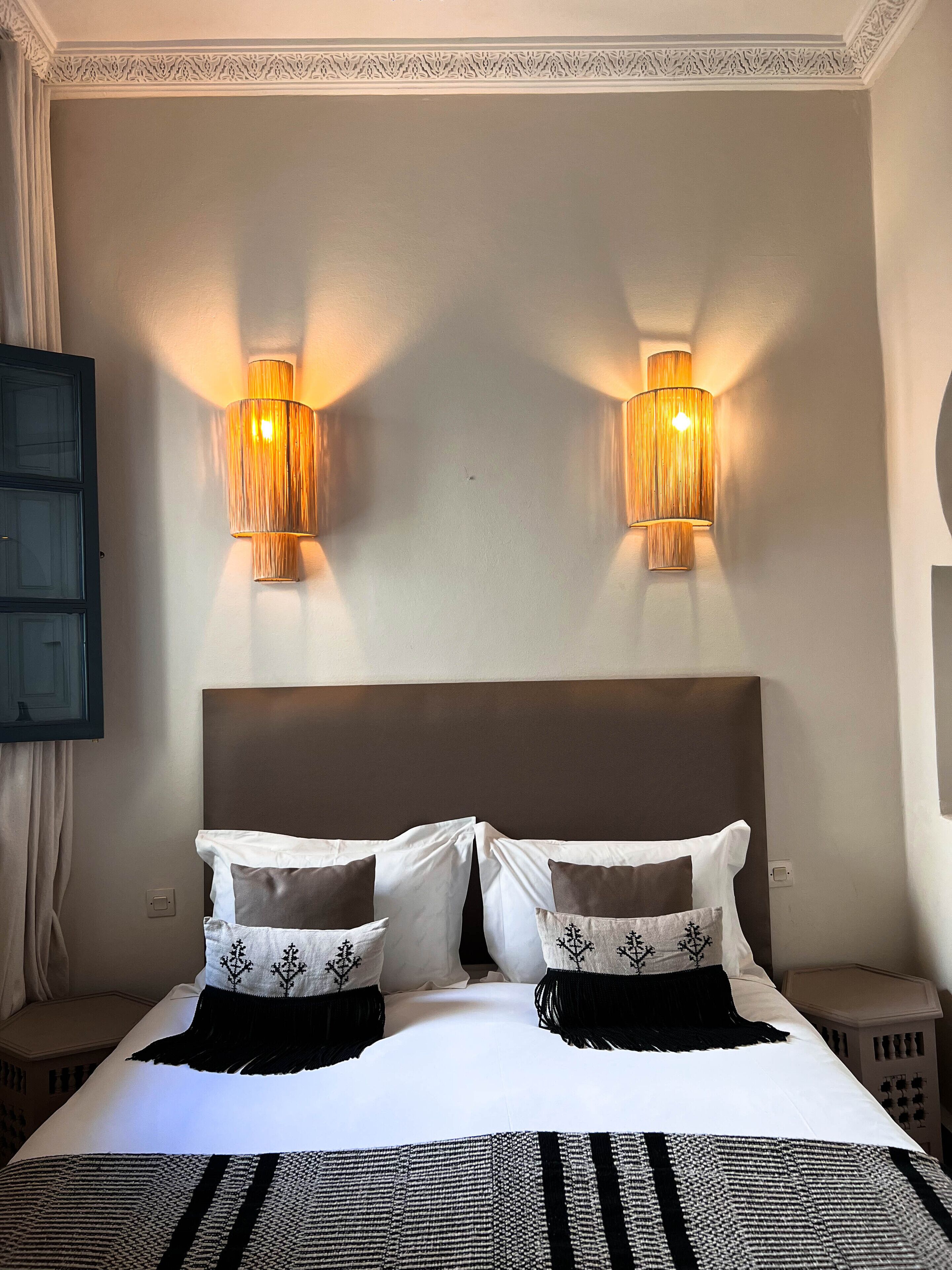 Cama bem arrumada com colcha estampada, duas luminárias na parede e cabeceira escura.