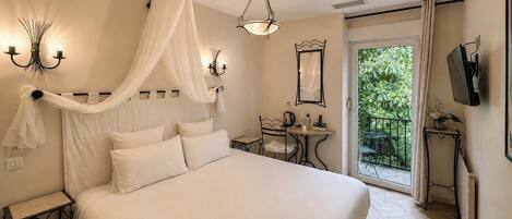 Quarto com cama, mesas de cabeceira, escrivaninha, cadeira, luminária pendente, iluminação no teto, televisão e vista para o verde lá fora.
