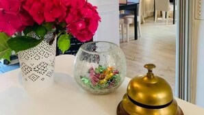 Quarto de hotel com uma mesa branca com um vaso de flores vermelhas, um enfeite de vidro com contas coloridas e um sino de latão.