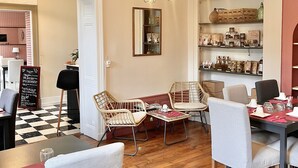 Uma sala de jantar com piso xadrez, mesas e cadeiras de madeira. Há luminárias pendentes e uma estante com diversos objetos.
