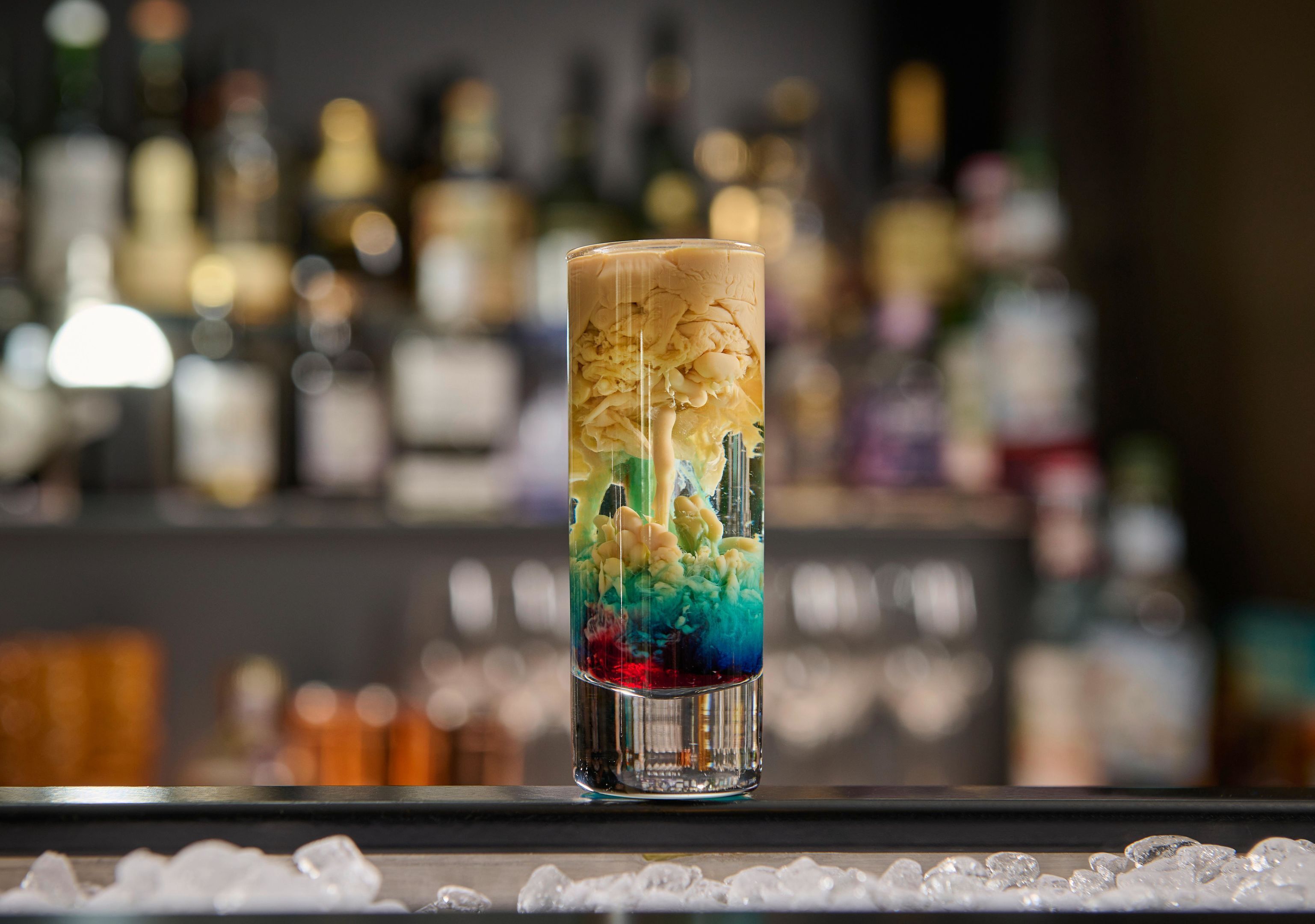 Um copo alto com uma bebida em camadas, cubos de gelo e um fundo desfocado de bar.