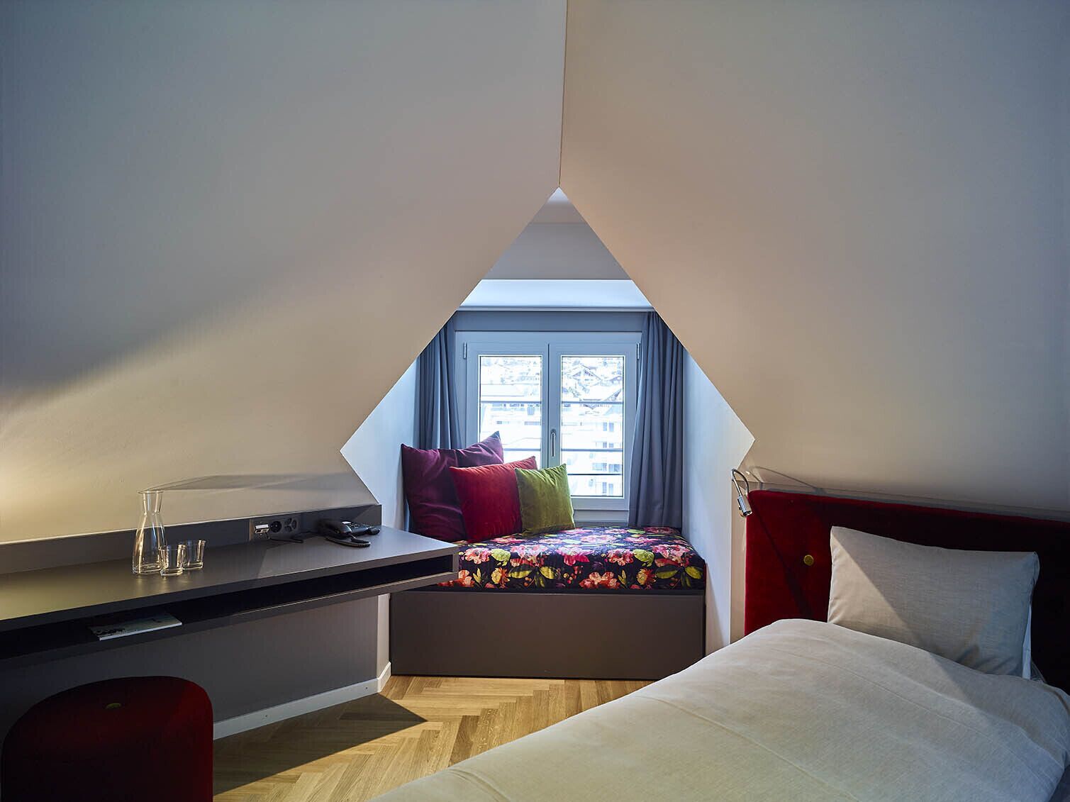 Quarto com teto inclinado, uma cama, uma escrivaninha e uma janela com cortinas.