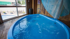 Uma grande piscina interior com um deck de madeira ao redor, vista para o exterior e um aviso de 'Não Fumar' na parede.