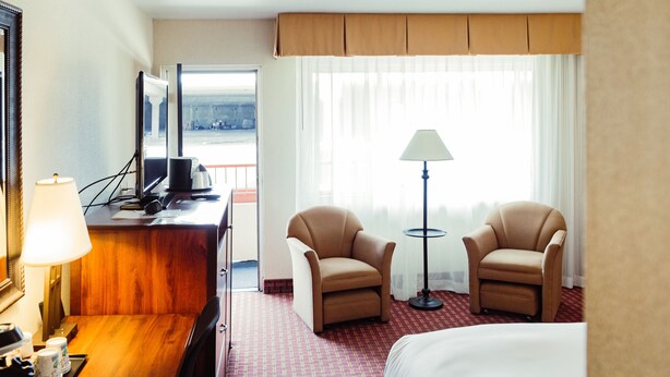 Quarto de hotel com uma cama, duas poltronas, uma escrivaninha com uma luminária, um computador e vista para a paisagem urbana pela janela.