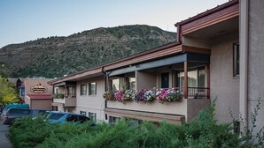 Um edifício com varanda decorada com flores, carros estacionados e montanhas ao fundo.