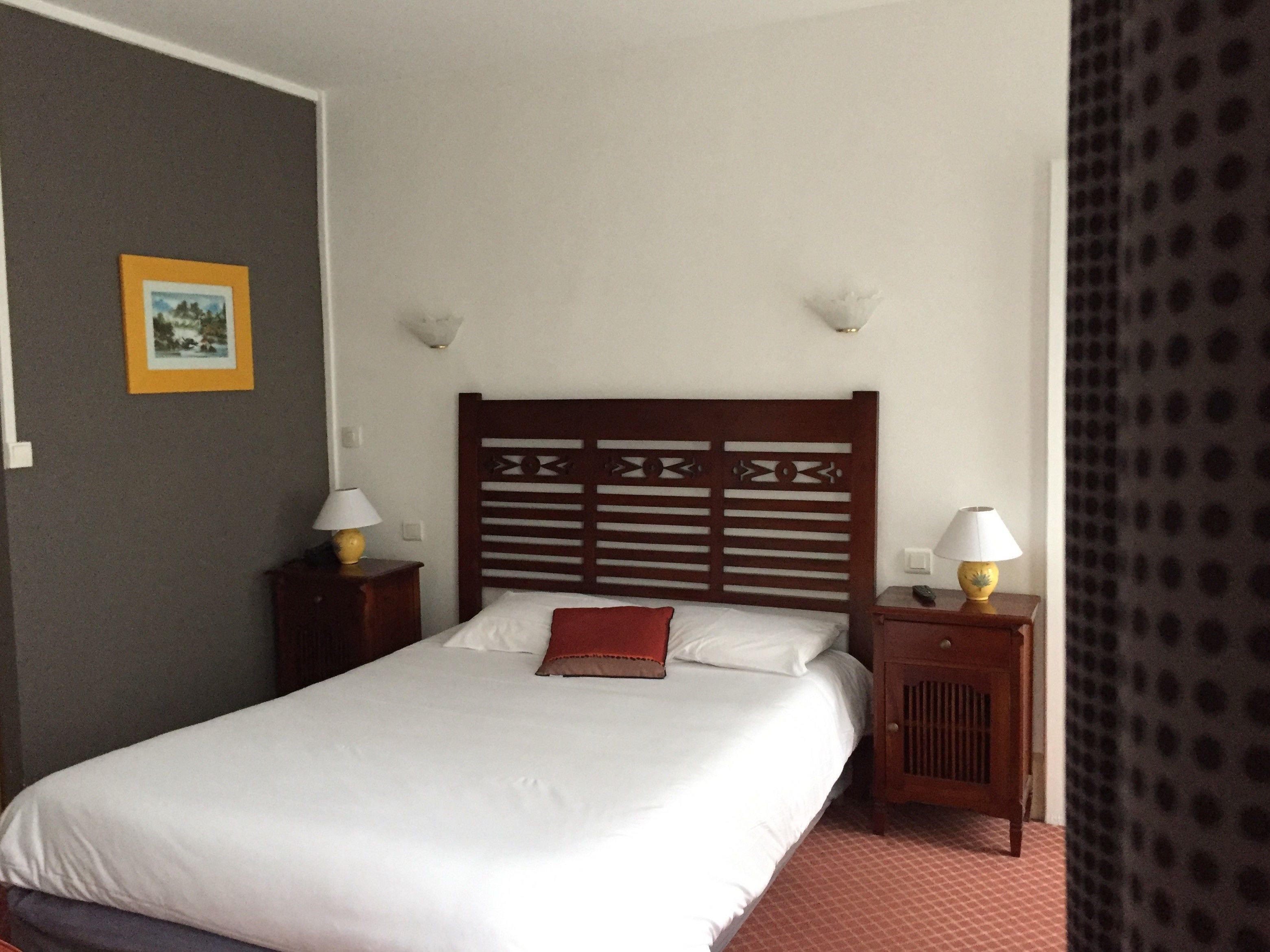 Cama bem arrumada com cabeceira de madeira, duas mesas de cabeceira com luminárias, um quadro na parede e cortina estampada.