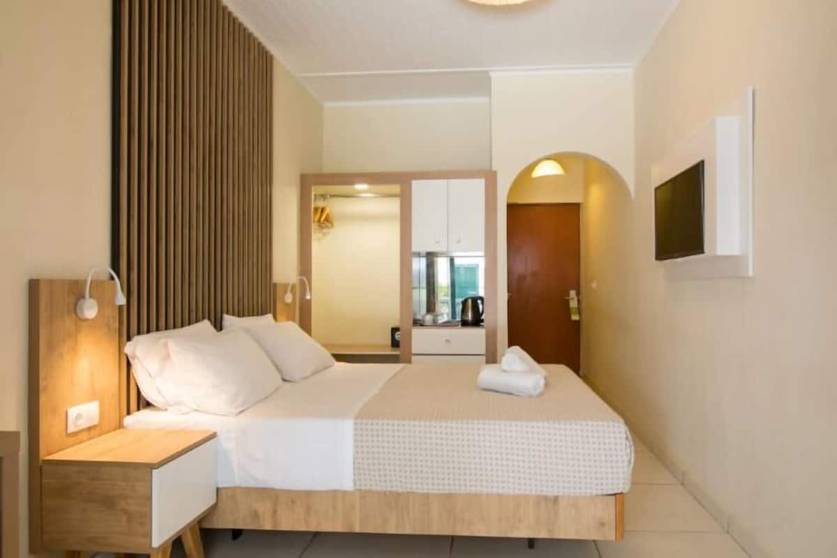 Quarto de hotel moderno com cabeceira de madeira, mesa de cabeceira com abajur, cama grande com colcha branca, TV de tela plana na parede e uma pequena mesa com chaleira.