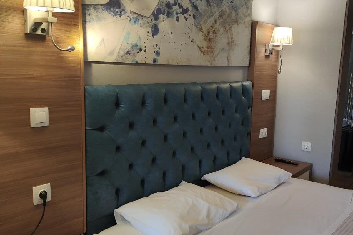 Quarto de hotel com cabeceira estofada, duas mesas de cabeceira, um quadro abstrato grande na parede e duas luminárias fixadas na parede.