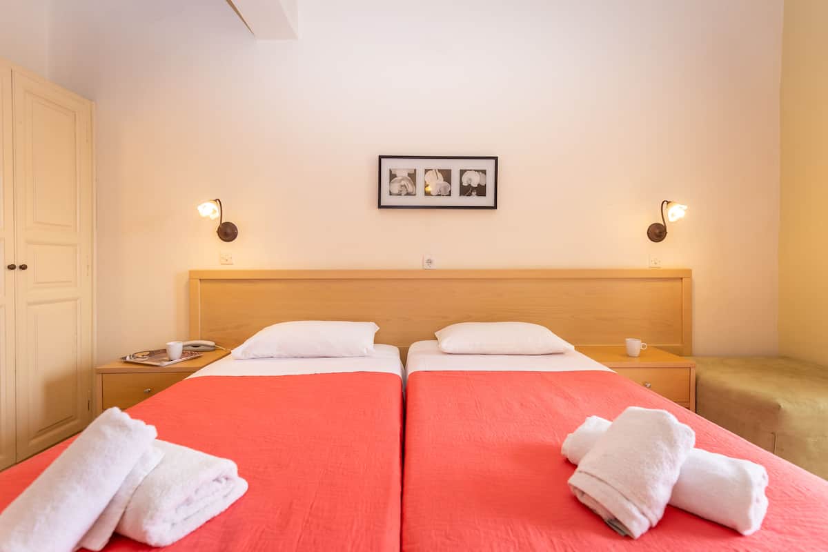 Quarto duplo com duas camas, cada uma com almofadas brancas e roupa de cama vermelha. Há duas mesas de cabeceira com abajures, um quadro emoldurado na parede e uma porta à esquerda.
