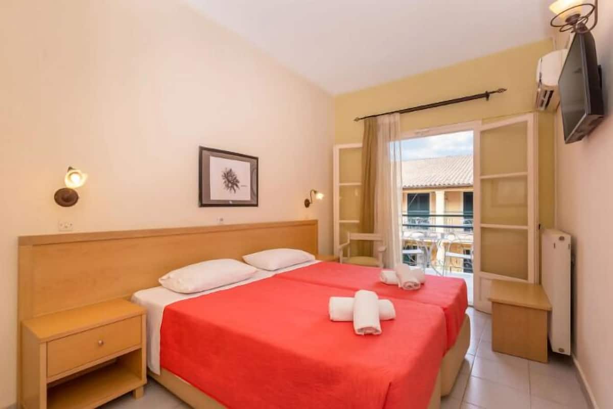 Quarto de hotel com cama de casal, mesa de cabeceira, televisão e varanda com vista.
