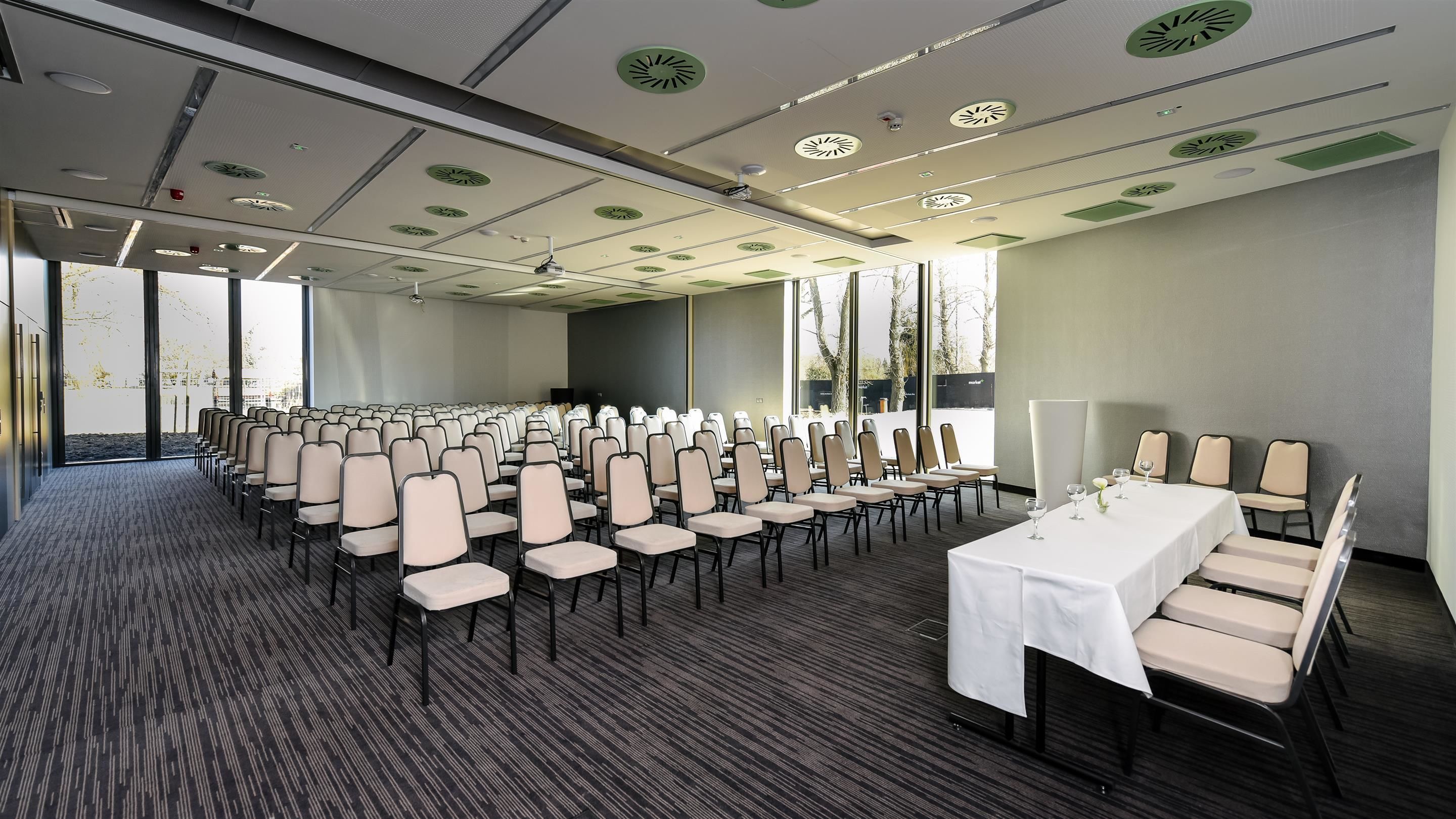 Uma sala de conferência com fileiras de cadeiras, uma mesa comprida com toalha branca e grandes janelas com vista para árvores.