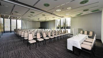Uma sala de conferência com fileiras de cadeiras, uma mesa comprida com toalha branca e grandes janelas com vista para árvores.