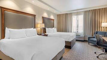 Quarto de hotel com duas camas grandes, uma escrivaninha com cadeira, uma luminária e uma janela ampla com cortinas.
