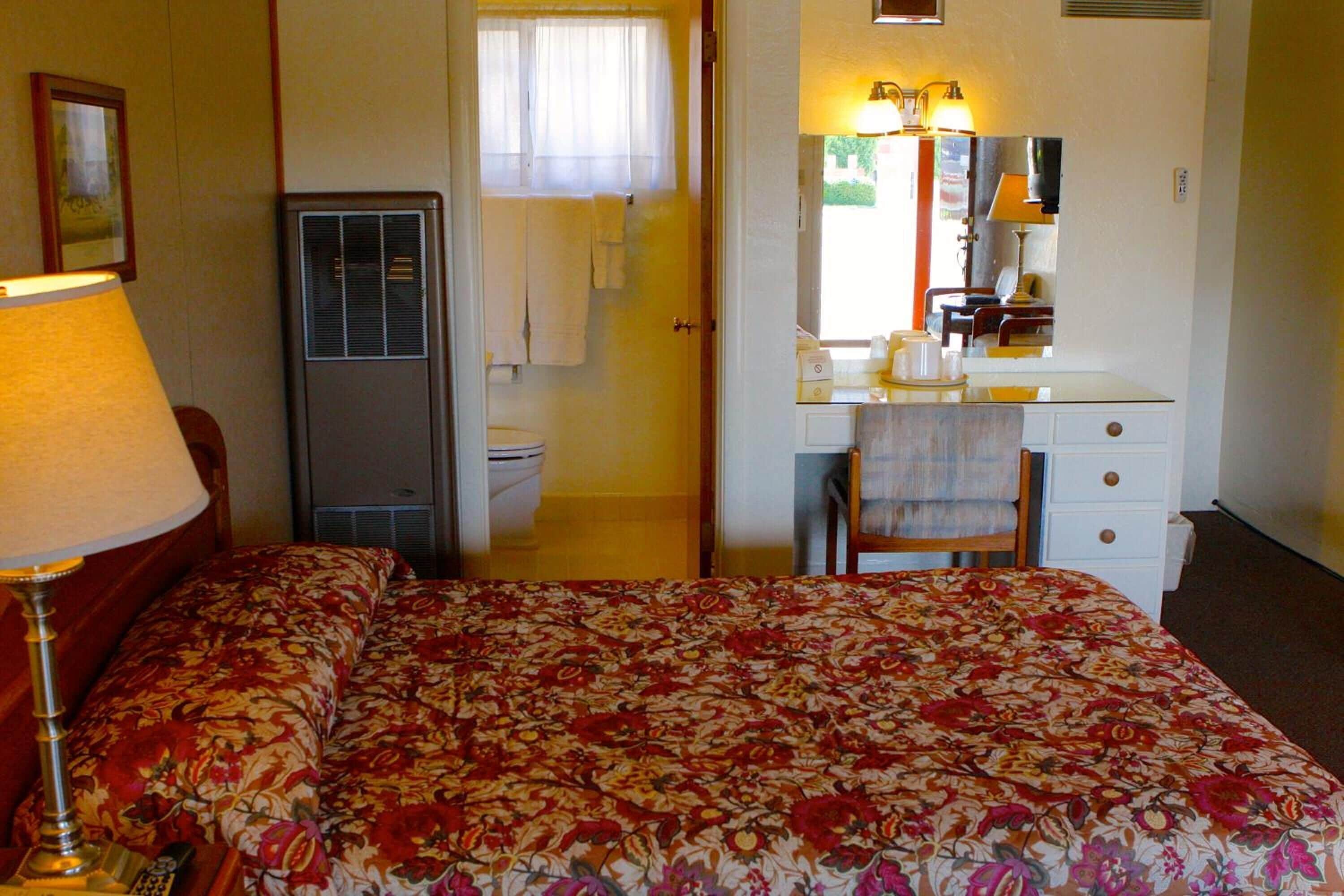 Quarto de hotel com uma cama, uma escrivaninha com cadeira, um espelho e um banheiro visível por uma porta aberta.