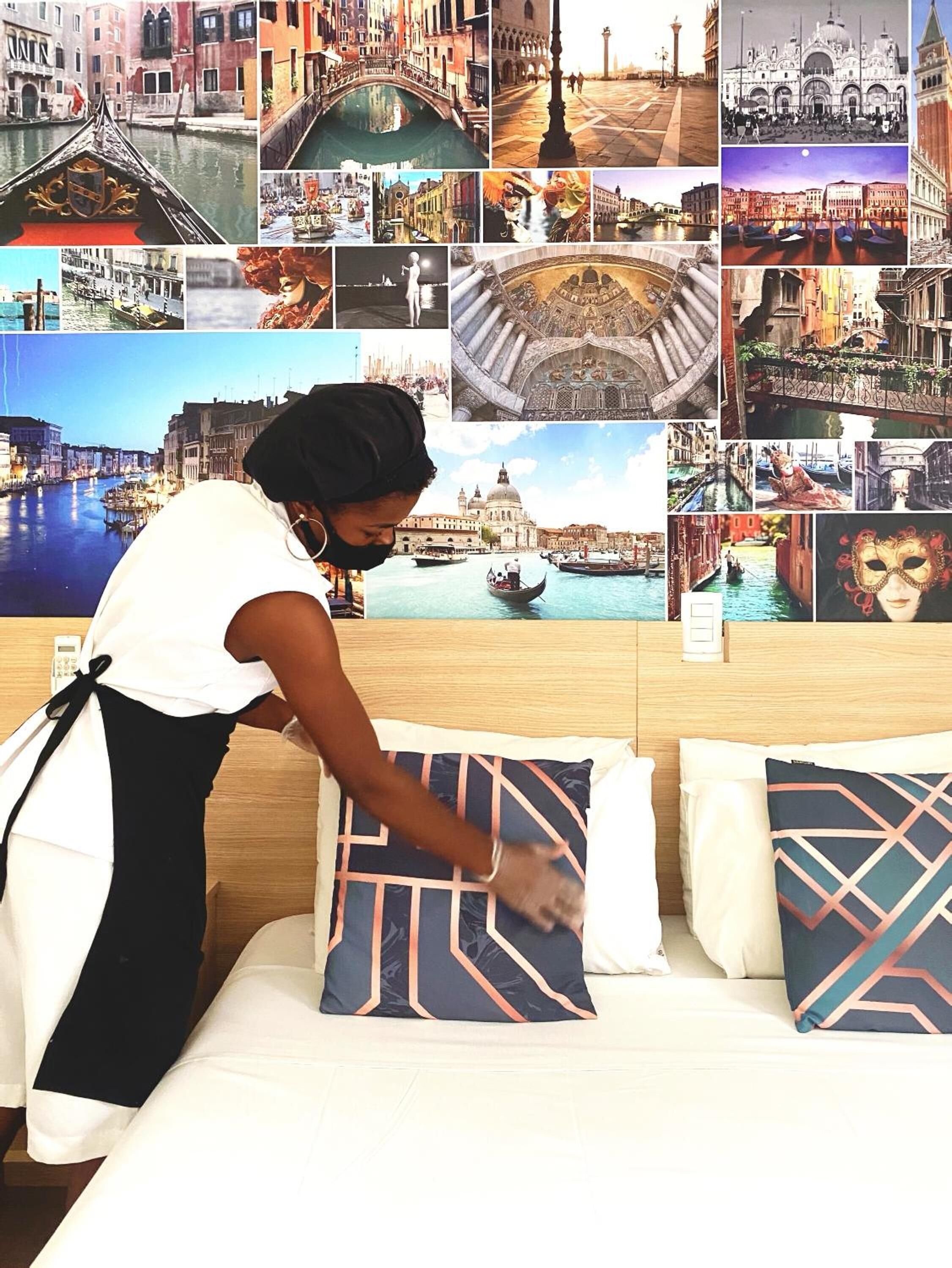 Quarto de hotel com cama, almofadas e parede decorada com diversas fotografias de viagens.